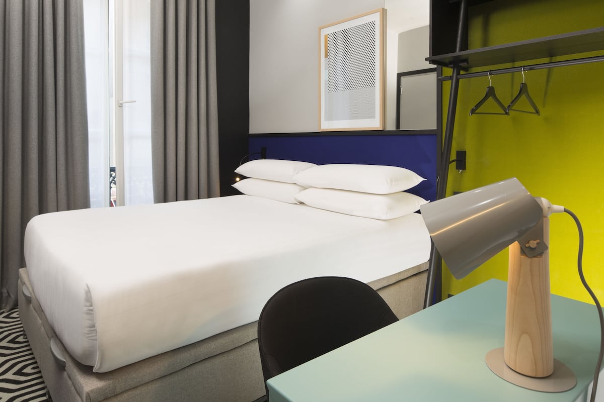 Quarto de hotel com cama, secretária com luminária, cadeira e janela com cortinas.