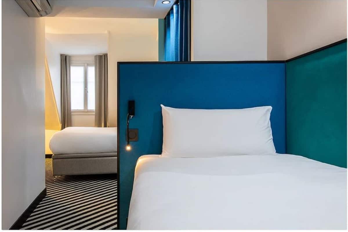 Um quarto de hotel com uma cama, mesa de cabeceira, abajur e janela com cortinas.