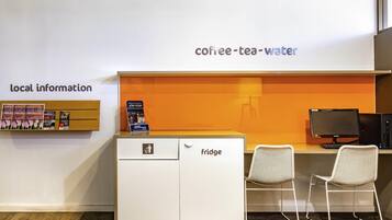 Bancada de recepção com sinalização para café, chá e água, uma geladeira e um monitor de computador.
