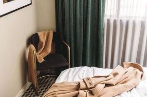 Quarto com cama, cadeira com manta, janela com cortinas e radiador.