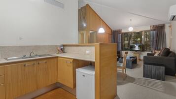 Cozinha compacta com pia, armários e uma pequena geladeira. Há uma área de refeições com mesa e cadeiras, e um sofá. O quarto tem uma janela com cortinas e uma luminária de teto.