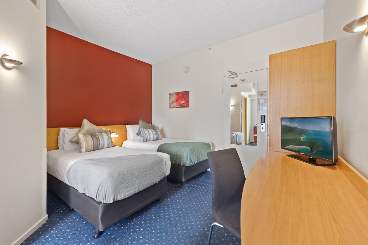 Quarto de hotel com duas camas, uma escrivaninha, uma televisão e um espelho.