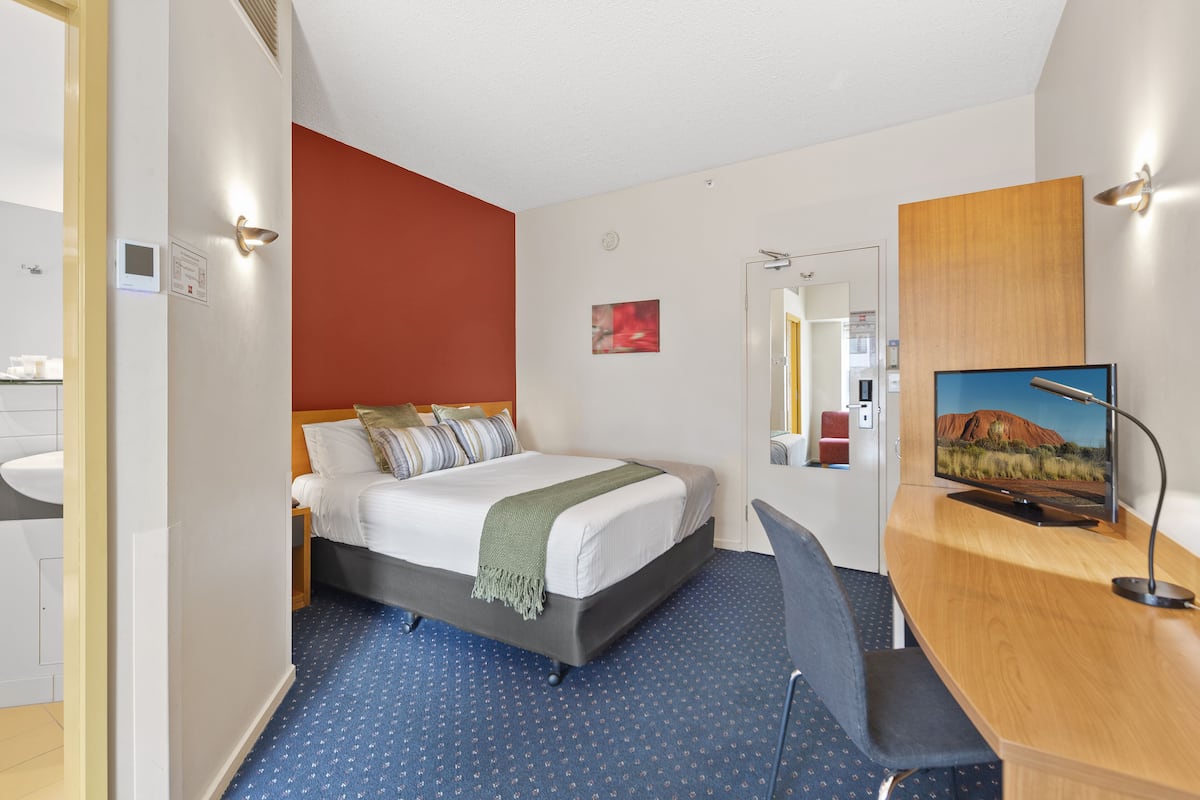 Quarto de hotel com cama, escrivaninha, cadeira, televisão e banheiro.