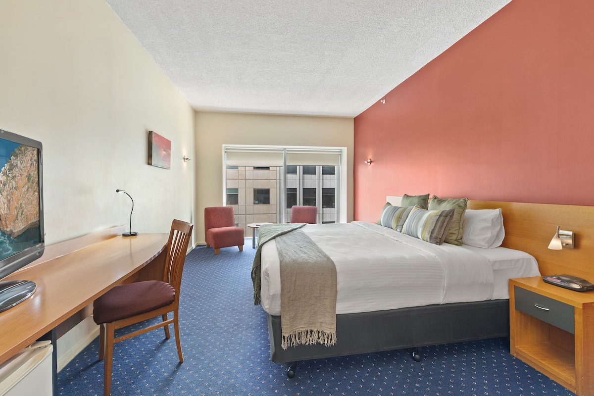 Quarto de hotel com cama, escrivaninha, cadeira e vista para o exterior.