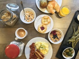Mesa de café da manhã com diversos alimentos, incluindo croissants, ovos mexidos, bacon e um copo de suco de laranja.