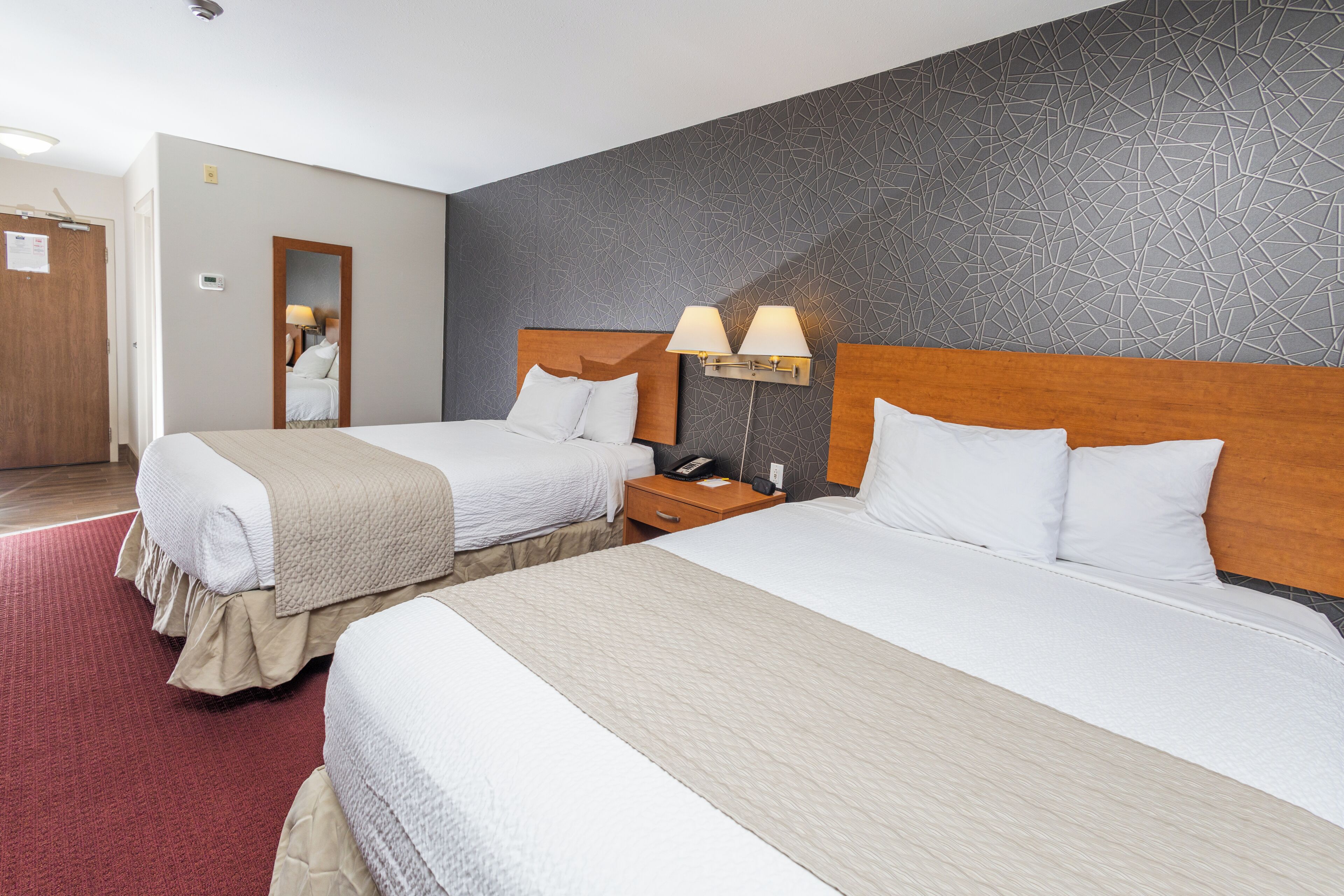 Quarto de hotel com duas camas, cabeceira de madeira, mesa de cabeceira com abajur e parede texturizada.