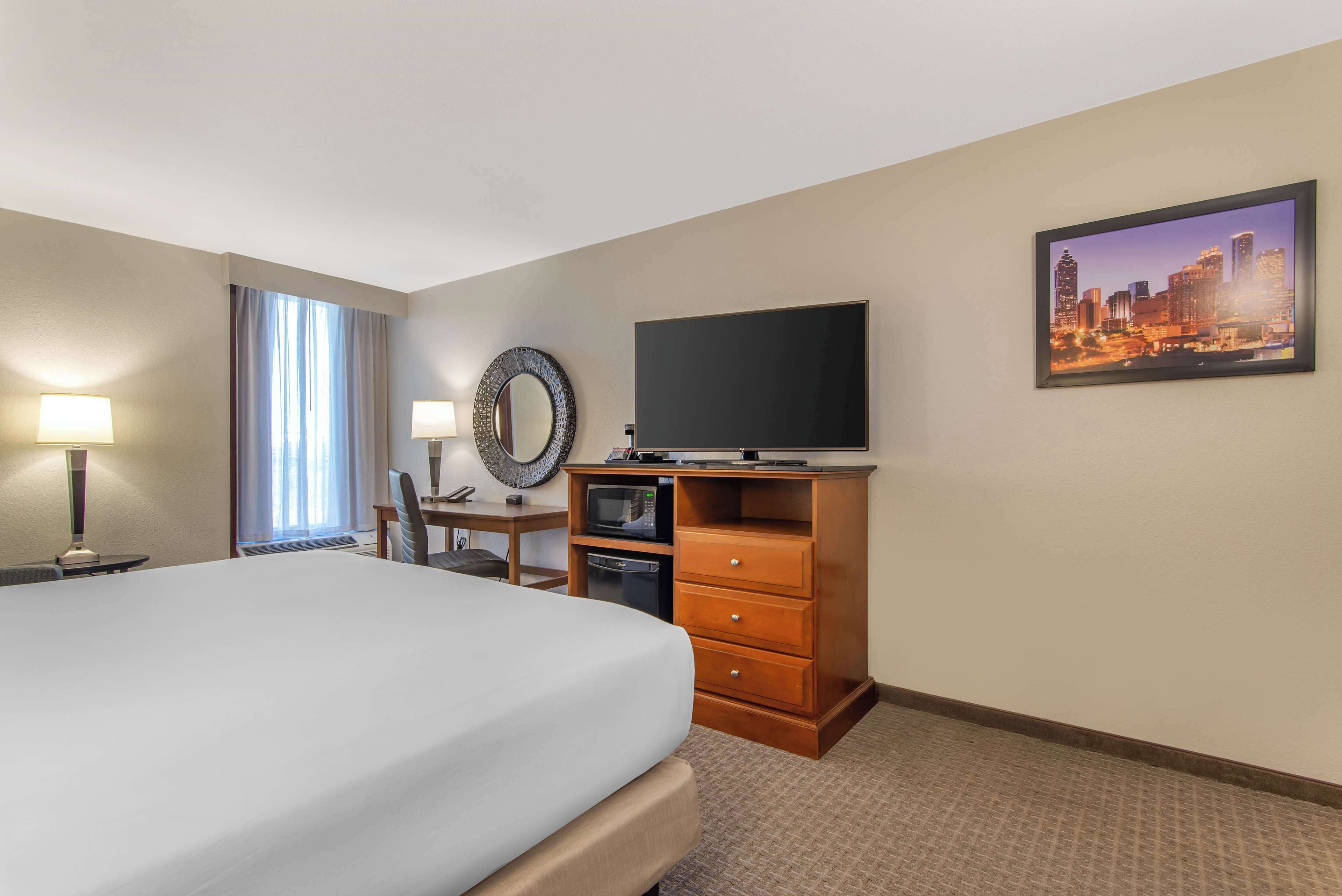 Quarto de hotel com uma cama grande, uma escrivaninha com cadeira, televisão, micro-ondas e uma janela ampla com cortinas.