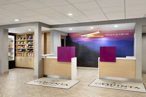 Uma área de recepção de hotel com design moderno, apresentando um balcão de atendimento, uma parede com um mural panorâmico e uma estante com lanches e bebidas.