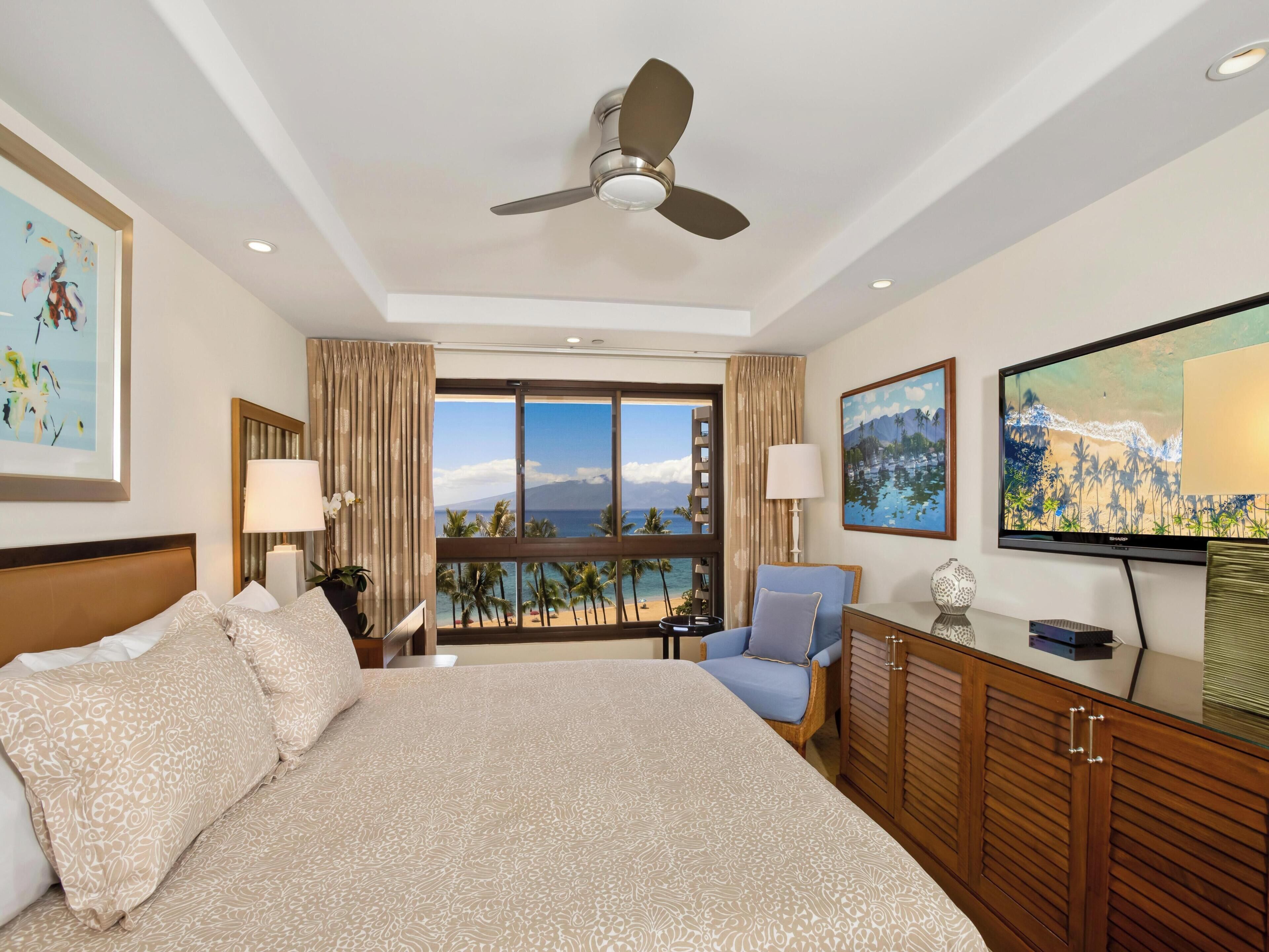 Quarto de hotel com uma cama grande, ventoinha no teto, televisão e vista para a praia.