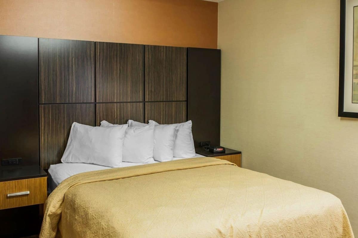 Quarto de hotel com cama, mesa de cabeceira, relógio fixado na parede e um quadro emoldurado.