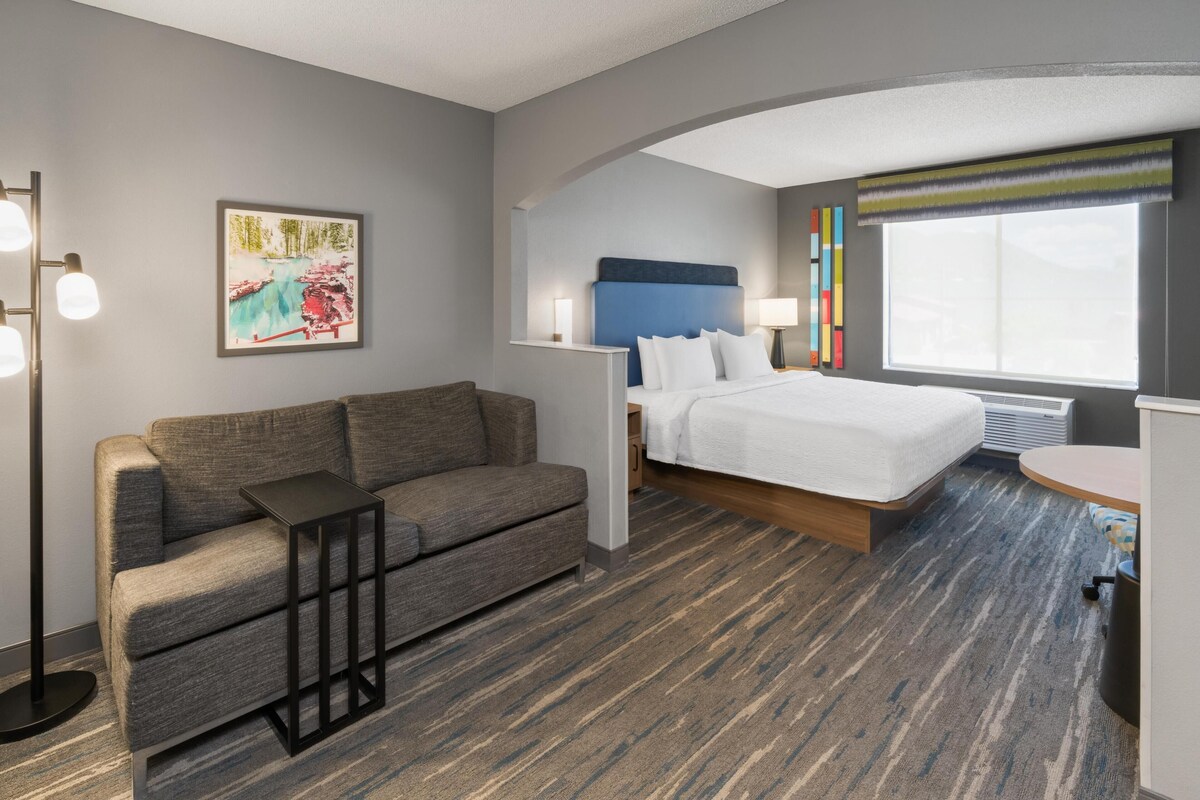 Quarto de hotel com cama, sofá, mesinha, um quadro e janela com vista.