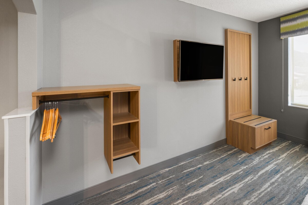Quarto com televisão de tela plana na parede, armário de madeira com gavetas e pequena escrivaninha com cadeira.