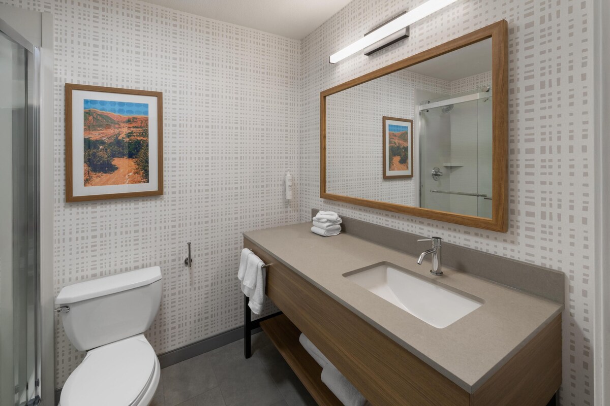Casa de banho com sanita, lavatório, espelho e um quadro emoldurado na parede.
