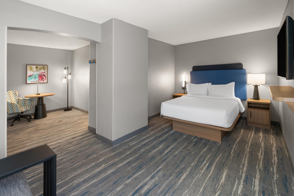 Quarto de hotel com cama, mesas de cabeceira, escrivaninha, cadeira e um quadro na parede.