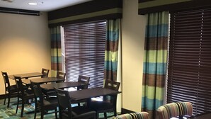 Uma sala de jantar de hotel com uma mesa comprida e várias cadeiras, um sofá com almofadas listradas e um tapete estampado.