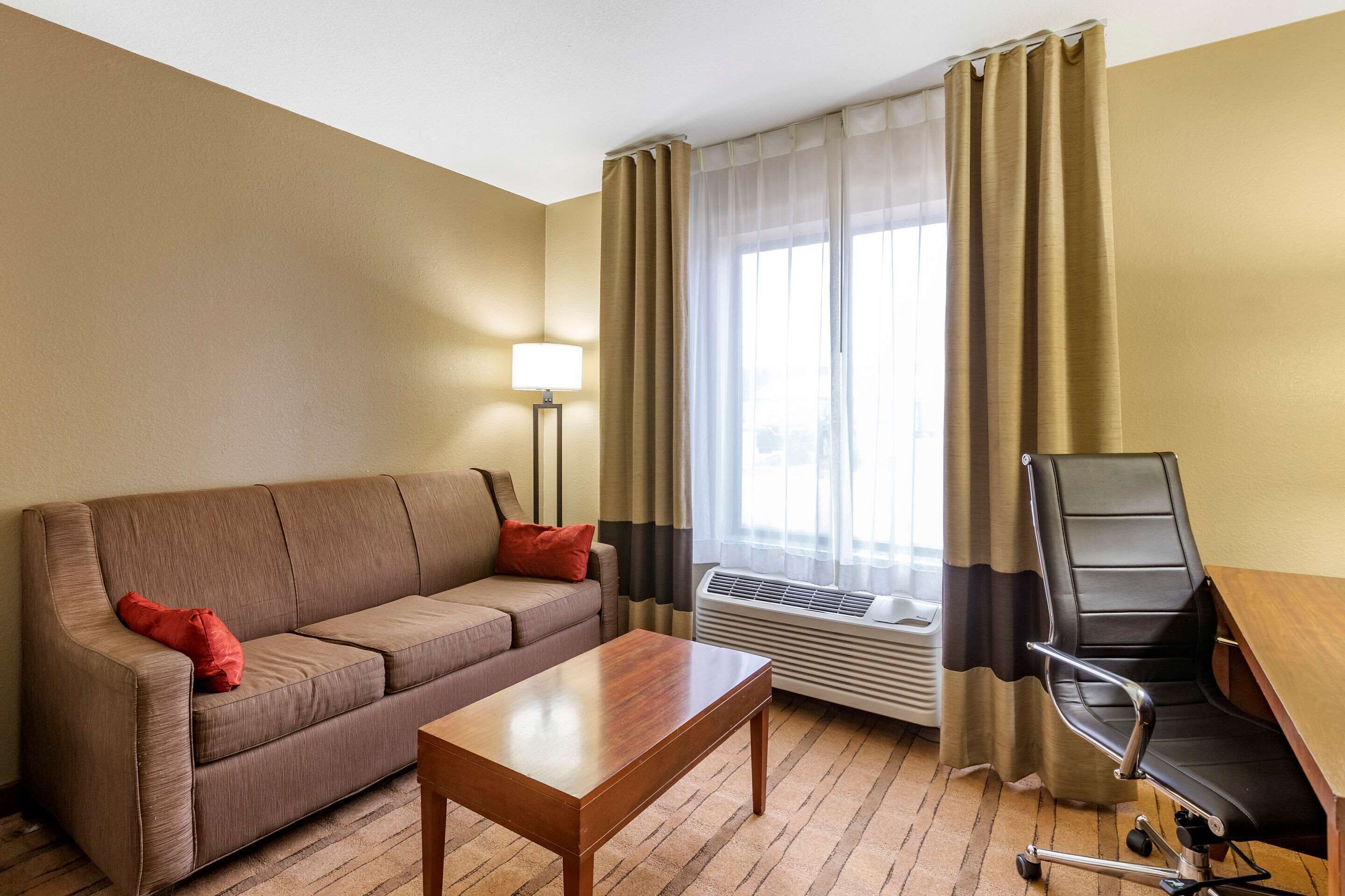 Quarto de hotel com sofá, mesa de centro de madeira, cadeira de escritório preta, abajur e janela com cortinas.