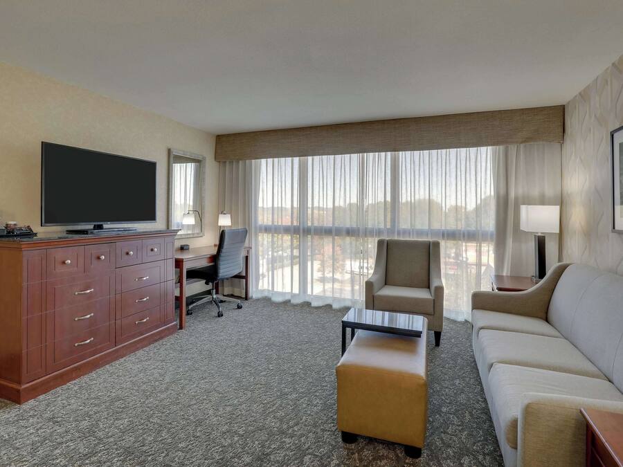 Quarto de hotel com uma televisão de tela plana grande, cômoda de madeira, escrivaninha com cadeira, sofá, pufe e uma janela grande com cortinas finas.