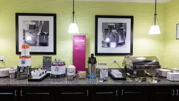 Uma estação de café com diversos equipamentos para preparo, um aviso divulgando um lanche e quadros nas paredes.