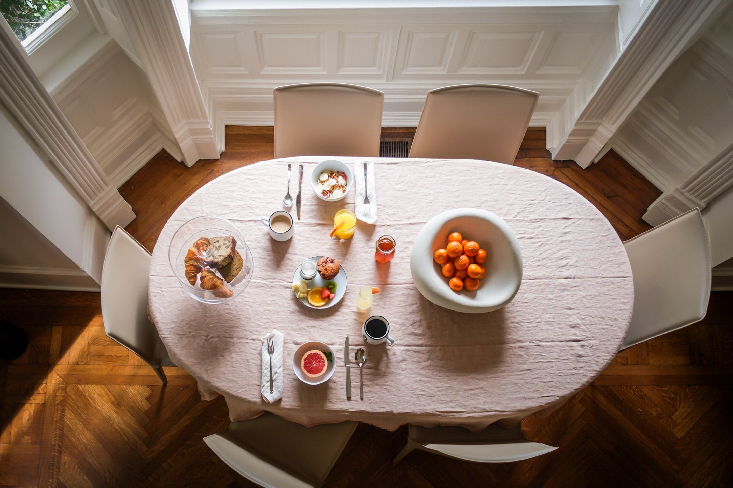 Mesa de jantar oval posta com itens de café da manhã, cercada por cadeiras em um ambiente com piso de madeira e paredes brancas.