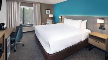Quarto de hotel com uma cama grande, uma escrivaninha, uma cadeira, uma luminária de mesa, um criado-mudo e uma janela com cortinas.