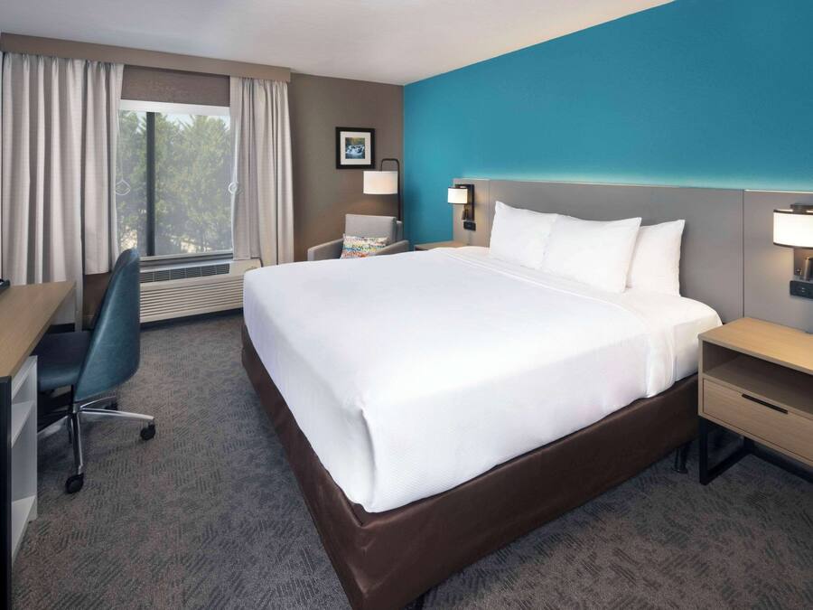 Quarto de hotel com uma cama grande, uma escrivaninha, uma cadeira, uma luminária de mesa, um criado-mudo e uma janela com cortinas.
