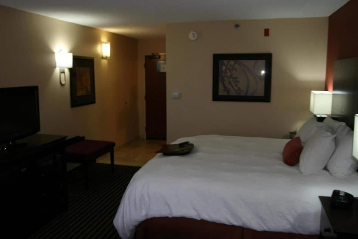 Quarto de hotel com cama, escrivaninha, cadeira, televisão e luminárias fixadas na parede.