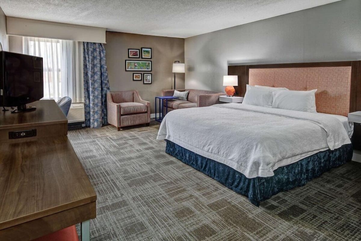 Quarto de hotel com uma cama, uma escrivaninha com computador, uma cadeira, um abajur e uma televisão.