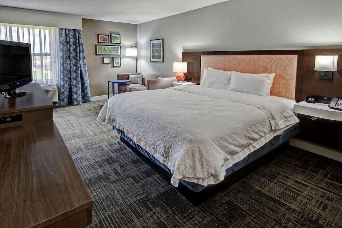 Quarto de hotel com uma cama grande, uma escrivaninha com televisão, uma cadeira, abajur e quadros nas paredes.