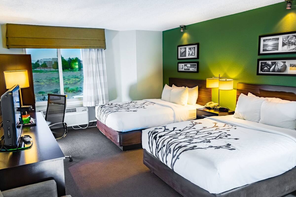 Quarto de hotel com duas camas, uma secretária com um computador, uma cadeira e uma janela com cortinas.