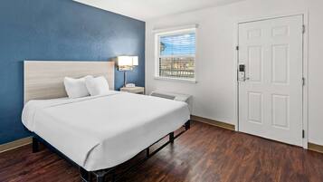 Quarto de hotel com cama, mesa de cabeceira, luminária, janela com persianas e porta.