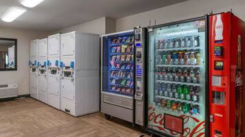 Quarto de hotel com uma fileira de mini-geladeiras brancas, uma máquina de vendas com a marca Coca-Cola e um espelho na parede.