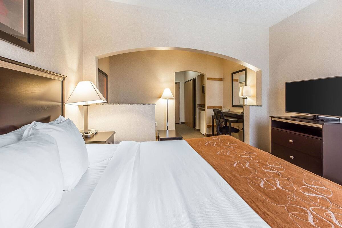 Quarto de hotel com uma cama, uma mesa, uma televisão e um banheiro visível por um arco.