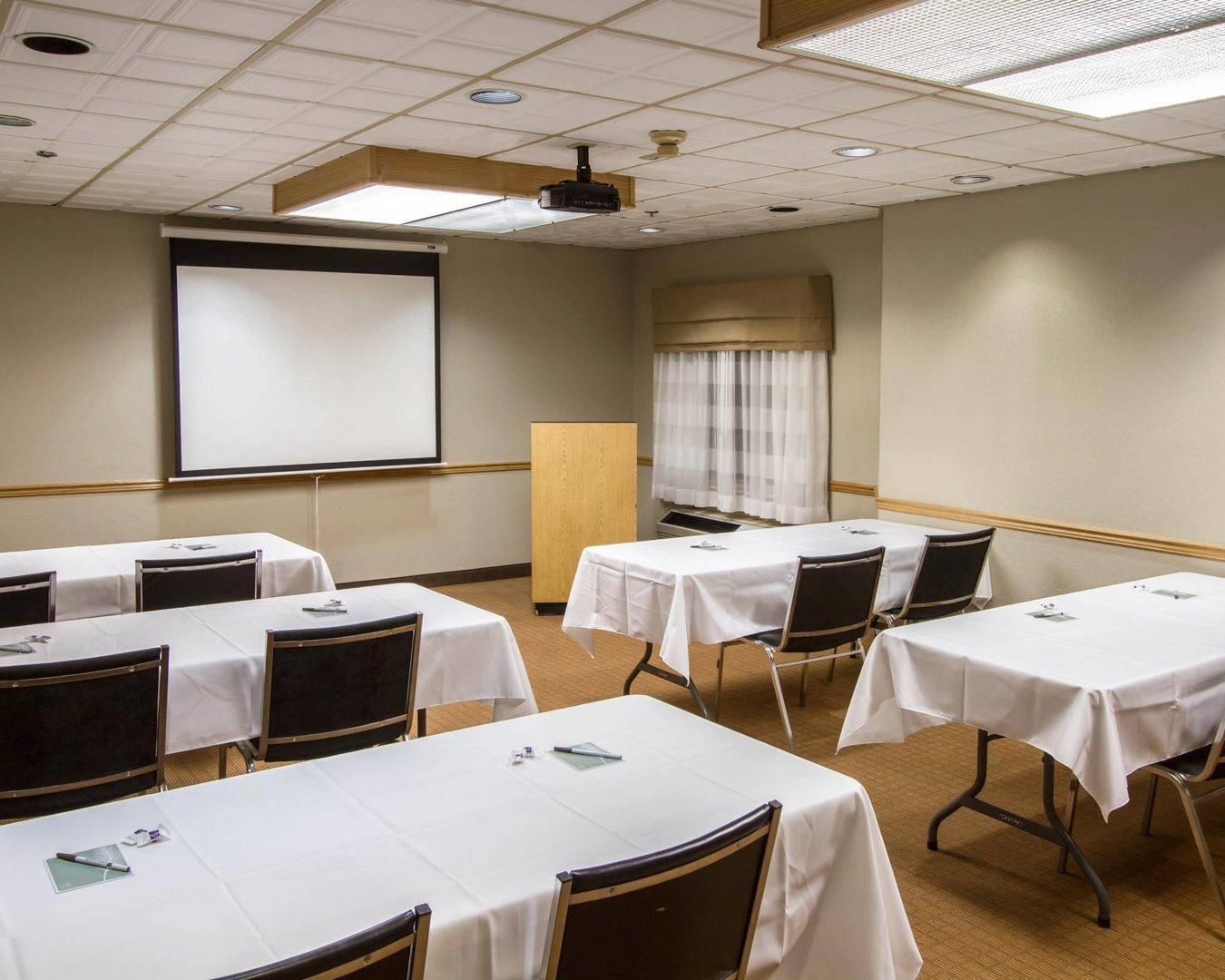 Uma sala de conferências com várias mesas, cadeiras, uma tela de projetor e um pódio.
