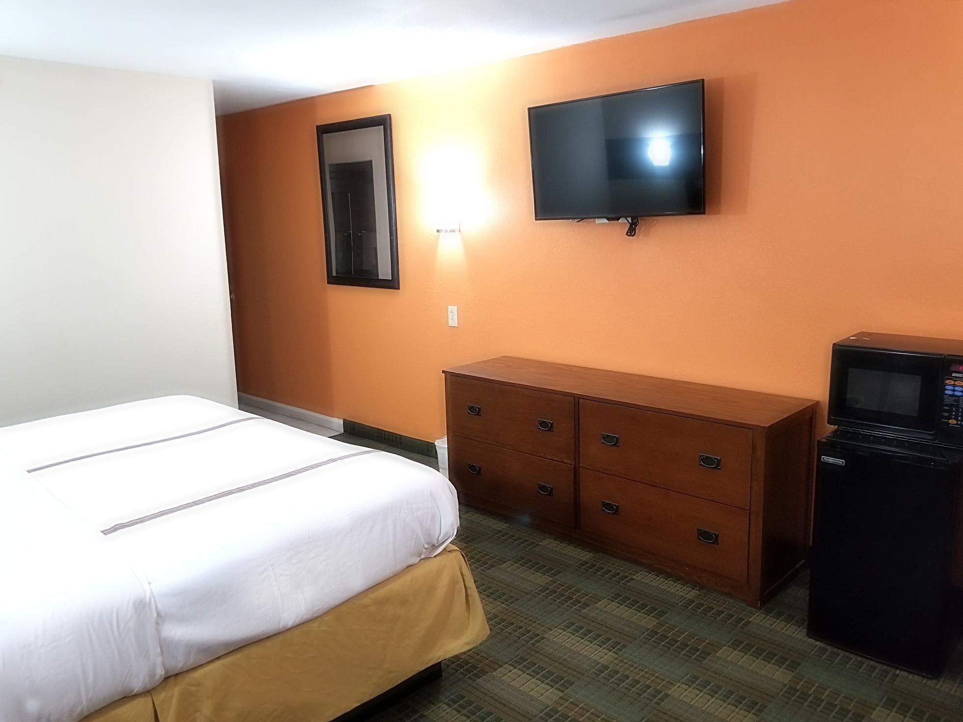 Quarto de hotel com uma cama, penteadeira de madeira, televisão na parede e micro-ondas sobre uma mesinha.