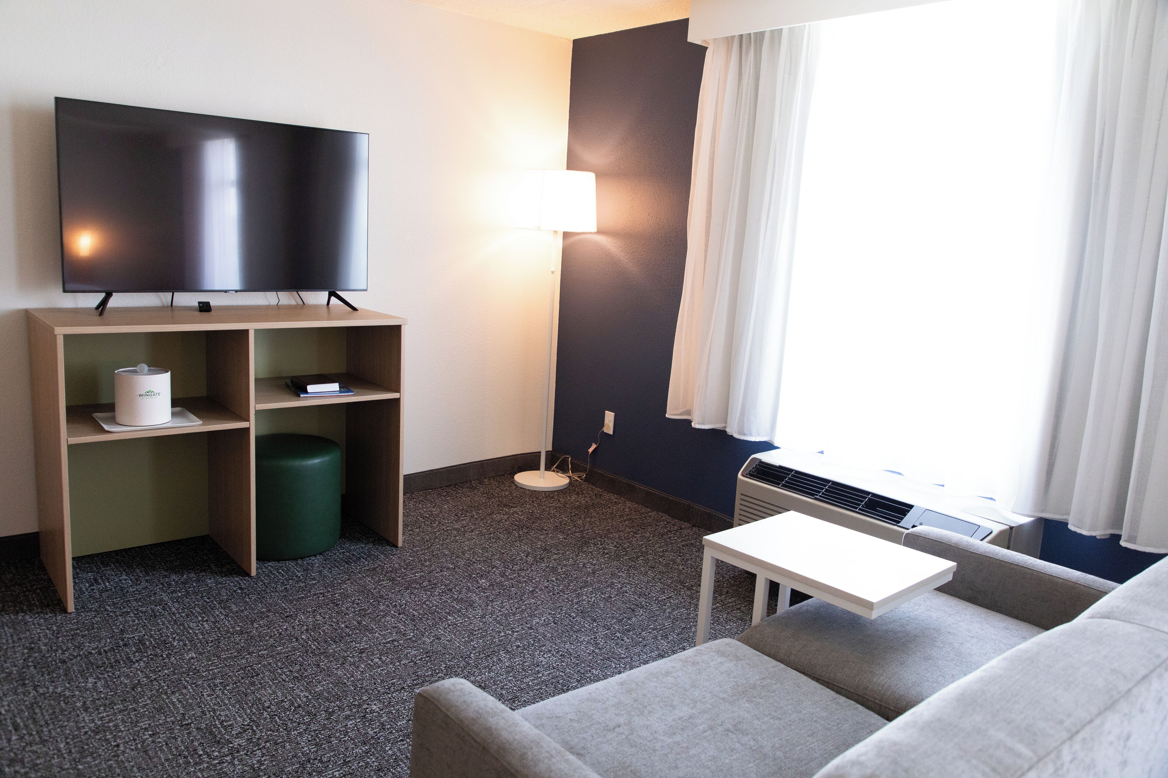 Quarto de hotel com televisão de tela plana em um suporte, uma mesinha, um sofá, uma lixeira e uma janela com cortinas.