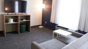 Quarto de hotel com televisão de tela plana em um suporte, uma mesinha, um sofá, uma lixeira e uma janela com cortinas.