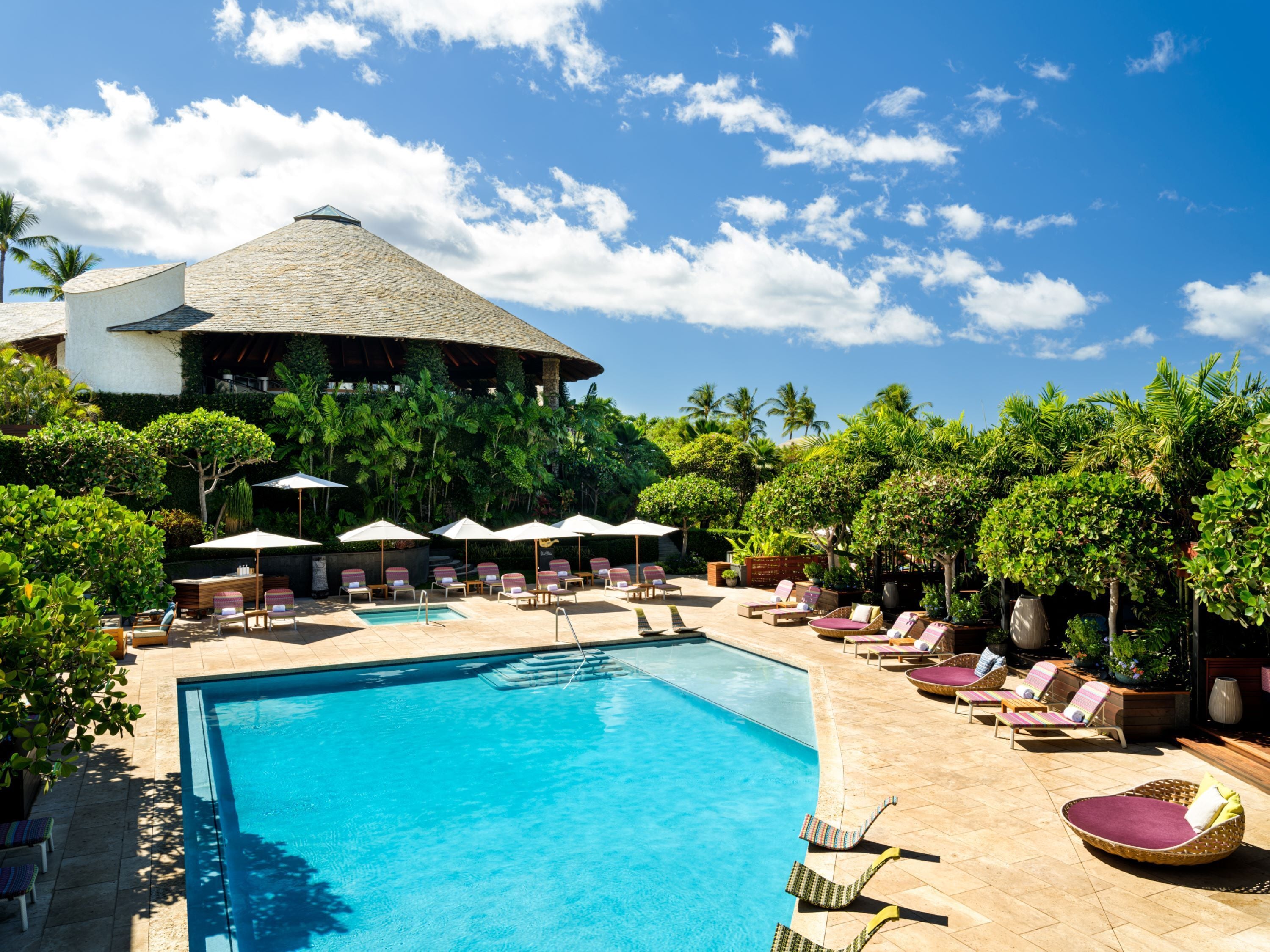 Piscina com espreguiçadeiras e guarda-sóis, cercada por vegetação exuberante e um edifício com telhado de palha.