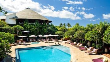 Piscina com espreguiçadeiras e guarda-sóis, cercada por vegetação exuberante e um edifício com telhado de palha.