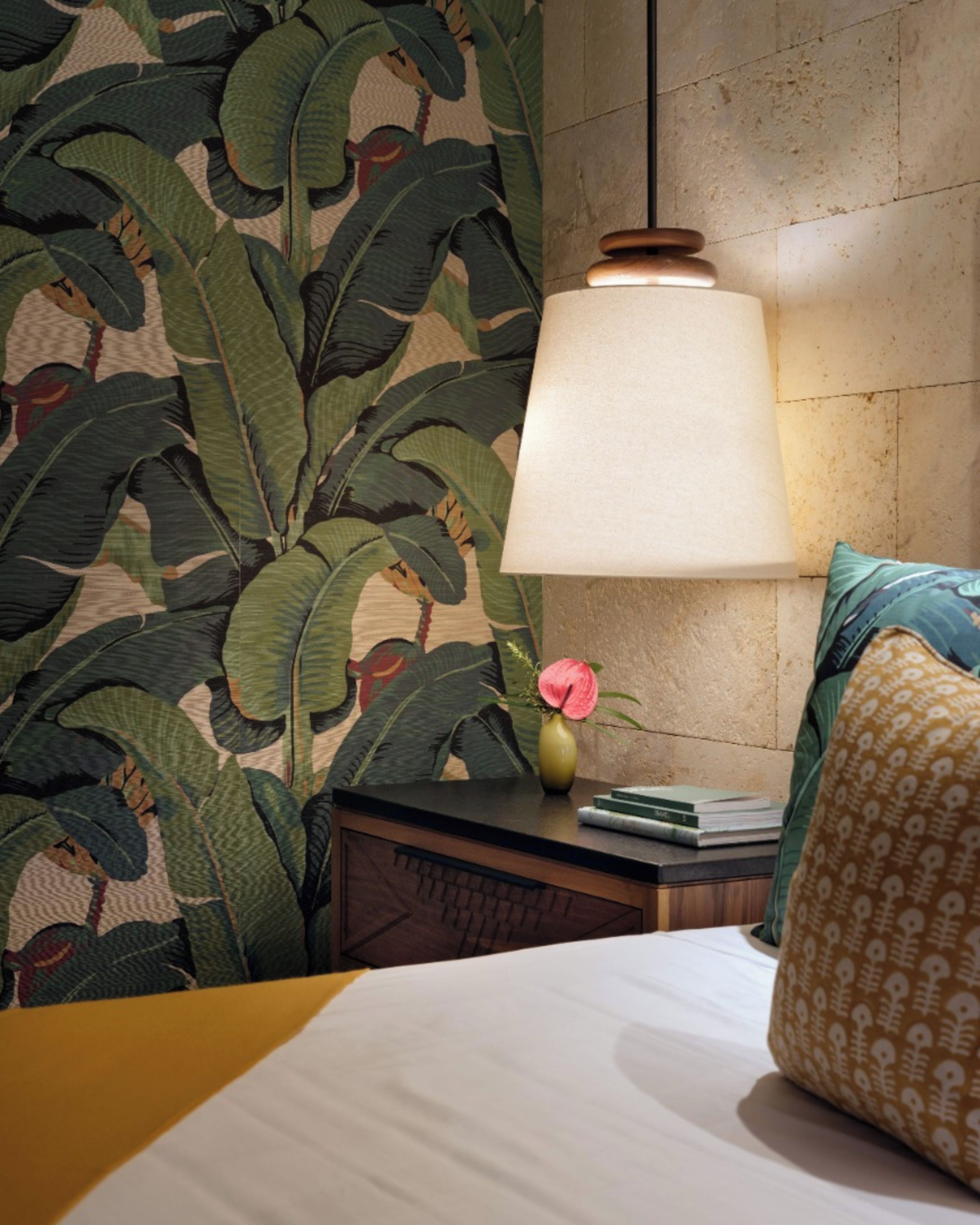 Quarto com cama, mesa de cabeceira com abajur, parede com padrão de folhas e travesseiro com estampa de caveira.