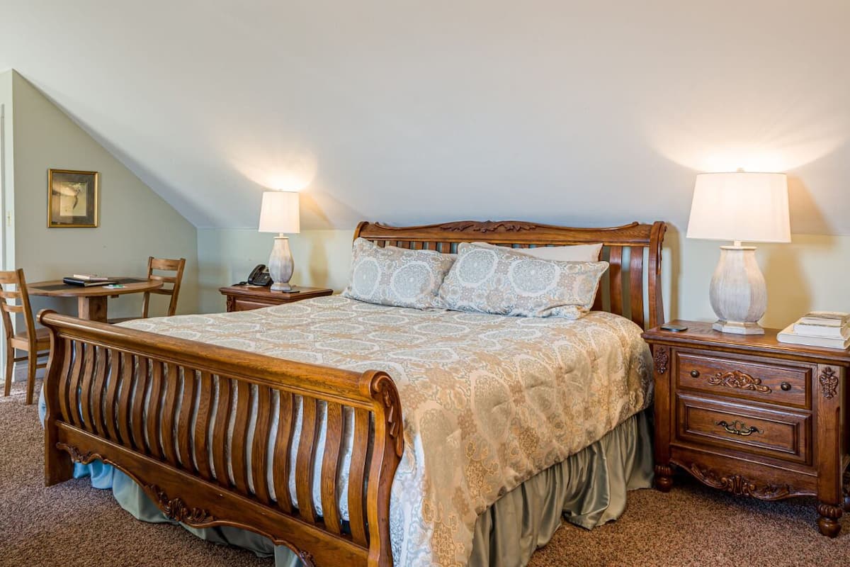 Quarto com cama de madeira, duas mesas de cabeceira com abajures, uma cadeira e uma escrivaninha.