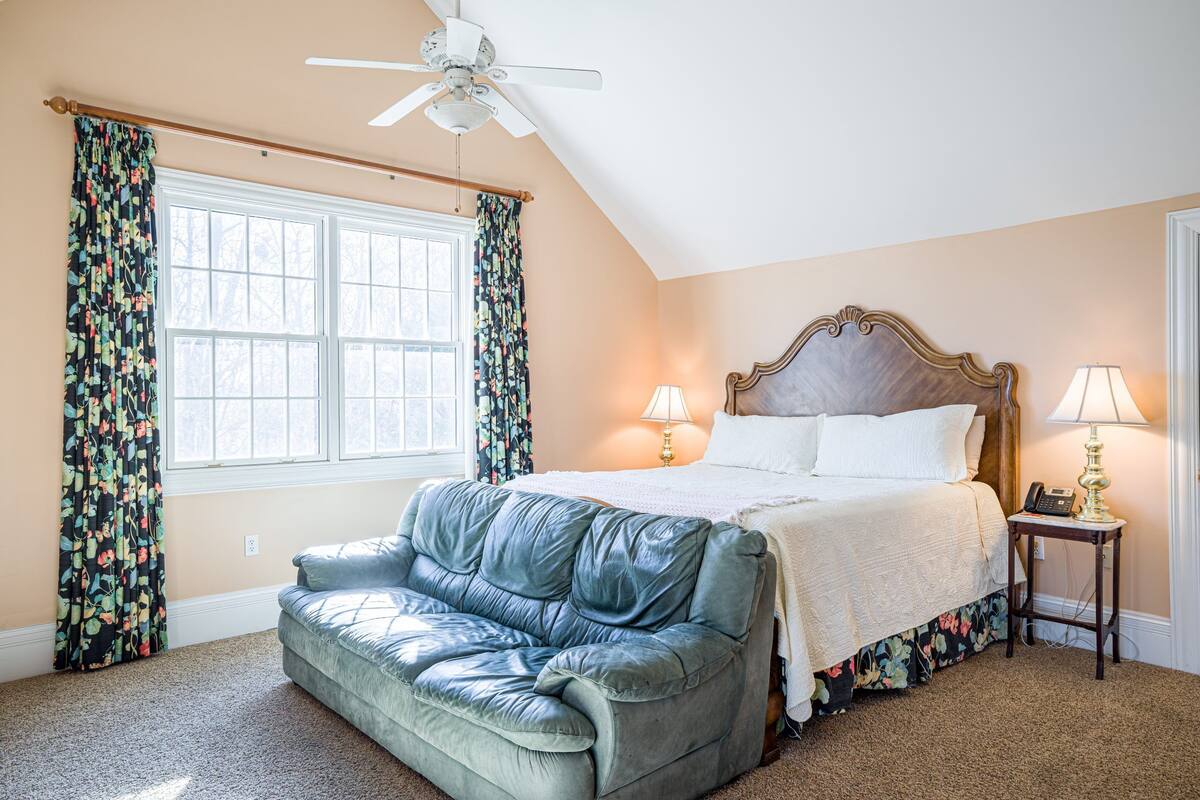 Um quarto com cama, duas luminárias de cabeceira, ventoinha no teto, cortina florida e sofá de couro.