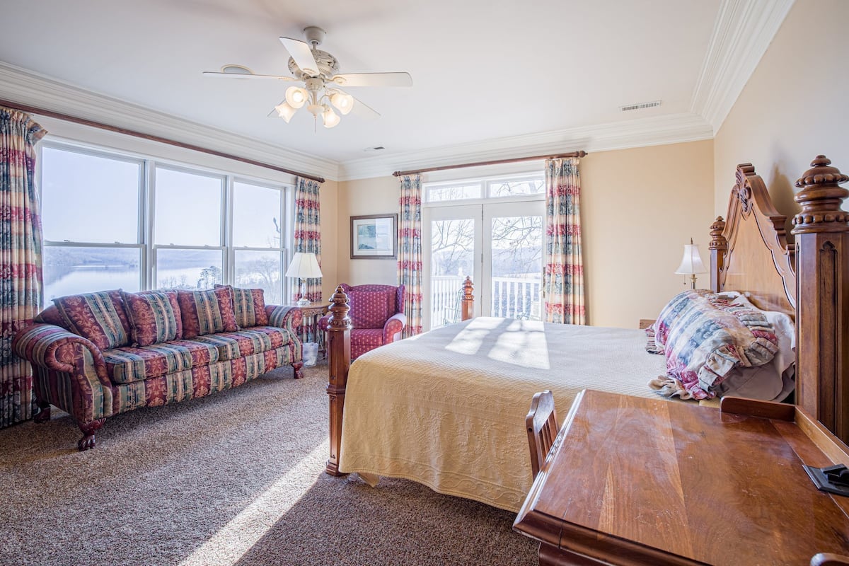 Quarto com uma cama grande, um sofá e uma cadeira. Há uma janela com cortinas e vista para o exterior.