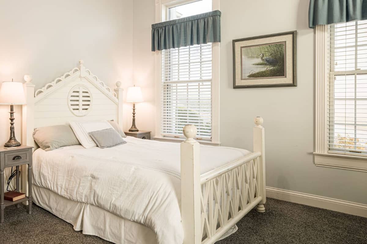 Quarto com cama branca, duas mesas de cabeceira com luminárias, uma janela com persianas e um quadro na parede.