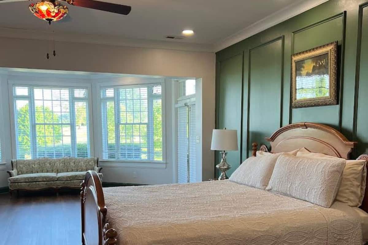 Quarto com cama grande, uma cadeira, mesa de cabeceira e janela com persianas.