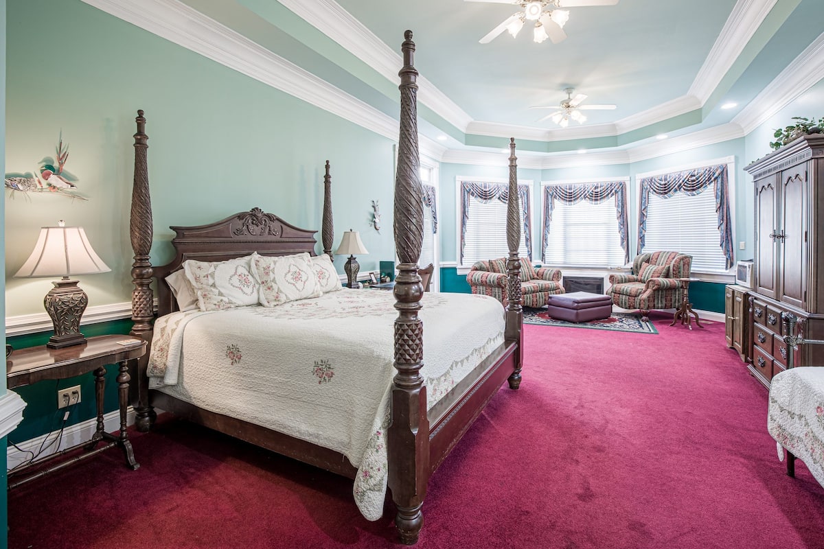 Um quarto espaçoso com cama de dossel, uma janela grande com cortinas, um ventilador de teto e uma cômoda de madeira.