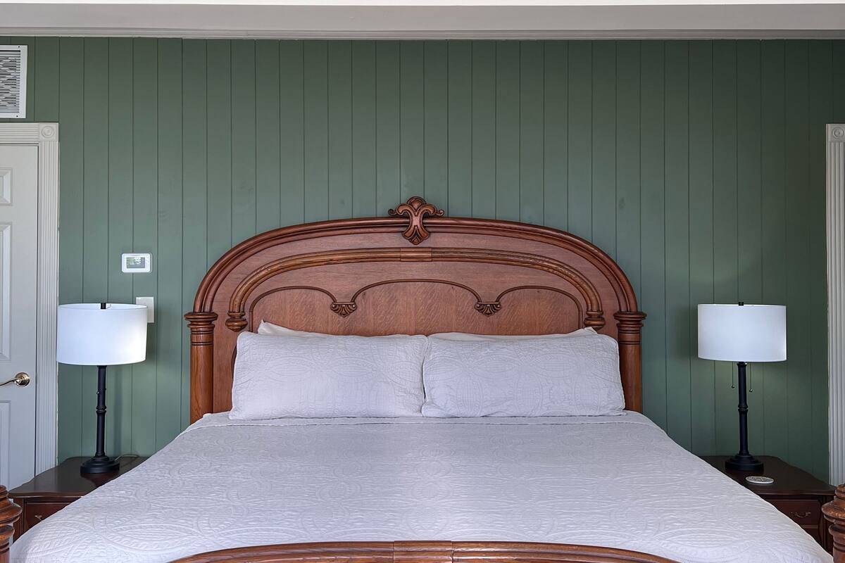 Uma cama com cabeceira de madeira, duas mesas de cabeceira com luminárias e paredes revestidas de verde.