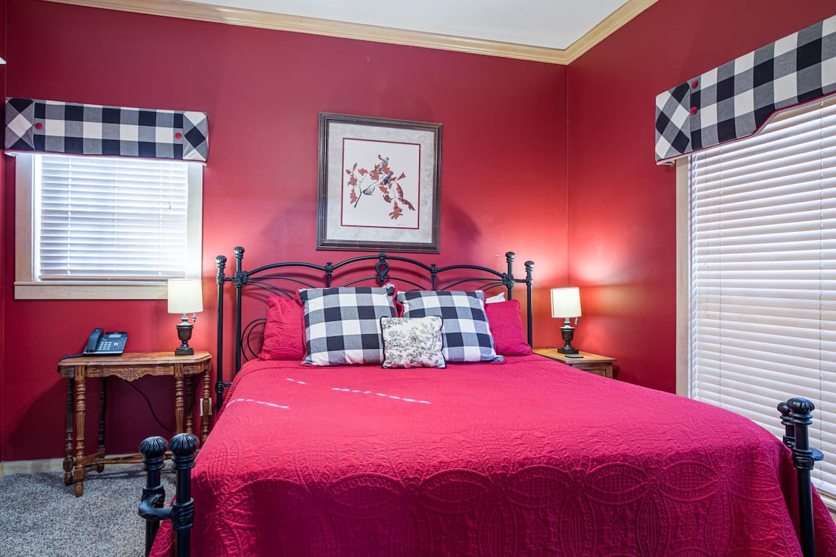 Quarto em tons de vermelho com colcha xadrez, duas mesas de cabeceira com abajures e um quadro na parede.