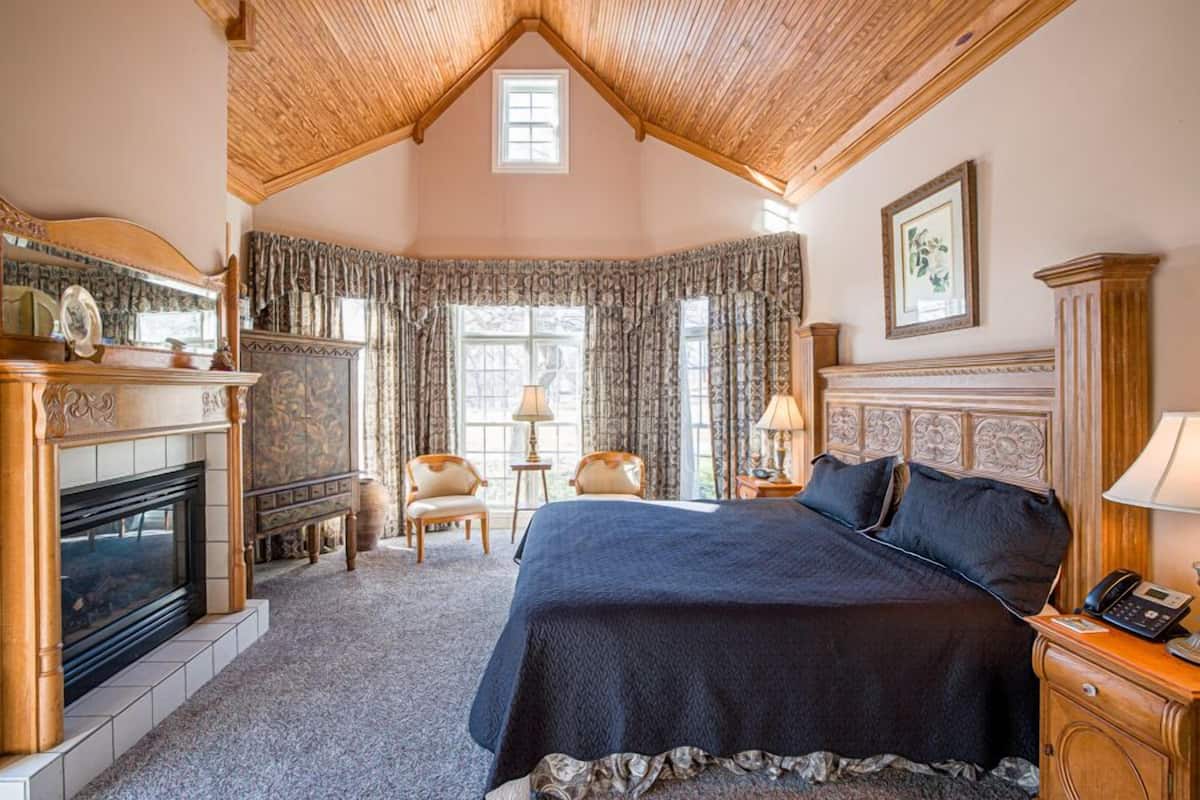 Quarto com lareira, uma cama grande, duas cadeiras, mesa de cabeceira e janela com cortinas.