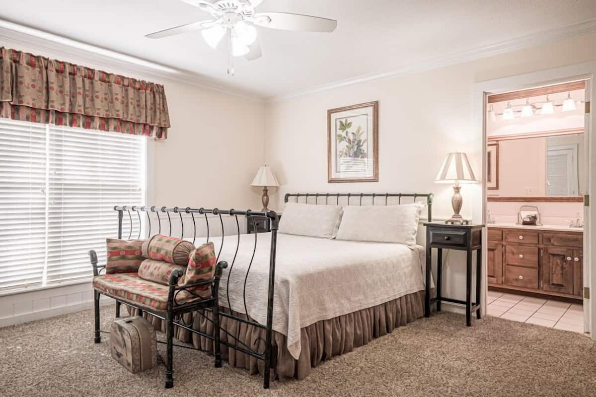 Quarto com cama, cadeira, mesa de cabeceira, luminária, ventilador de teto, janela com persianas, quadro na parede e área de cozinha com armários e bancada.