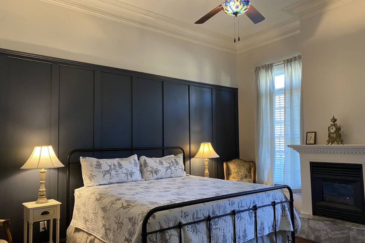 Quarto com uma cama grande, duas luminárias de cabeceira, lareira e ventilador de teto.